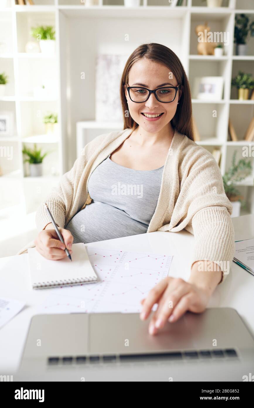 Smiling pretty pregnant lady in eyeglasses sitting at desk with graph ...