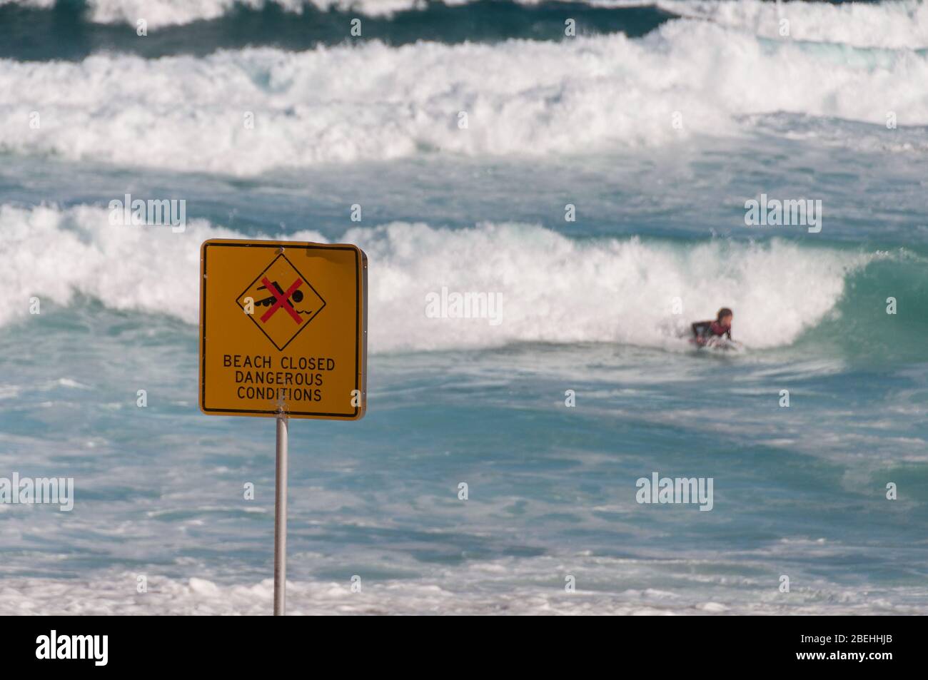 A warning sign of closed beach with person surfing rough waves on the ...