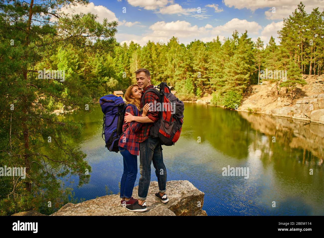 Cute couple with backpacks stand at rock near the lake at forest and ...