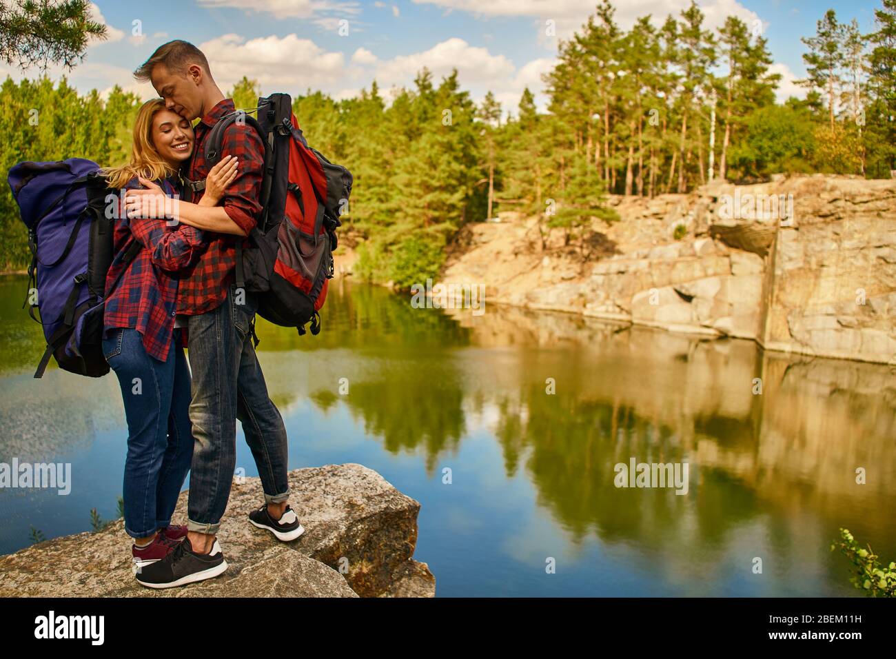 Cute couple with backpacks stand at rock near the lake at forest and ...