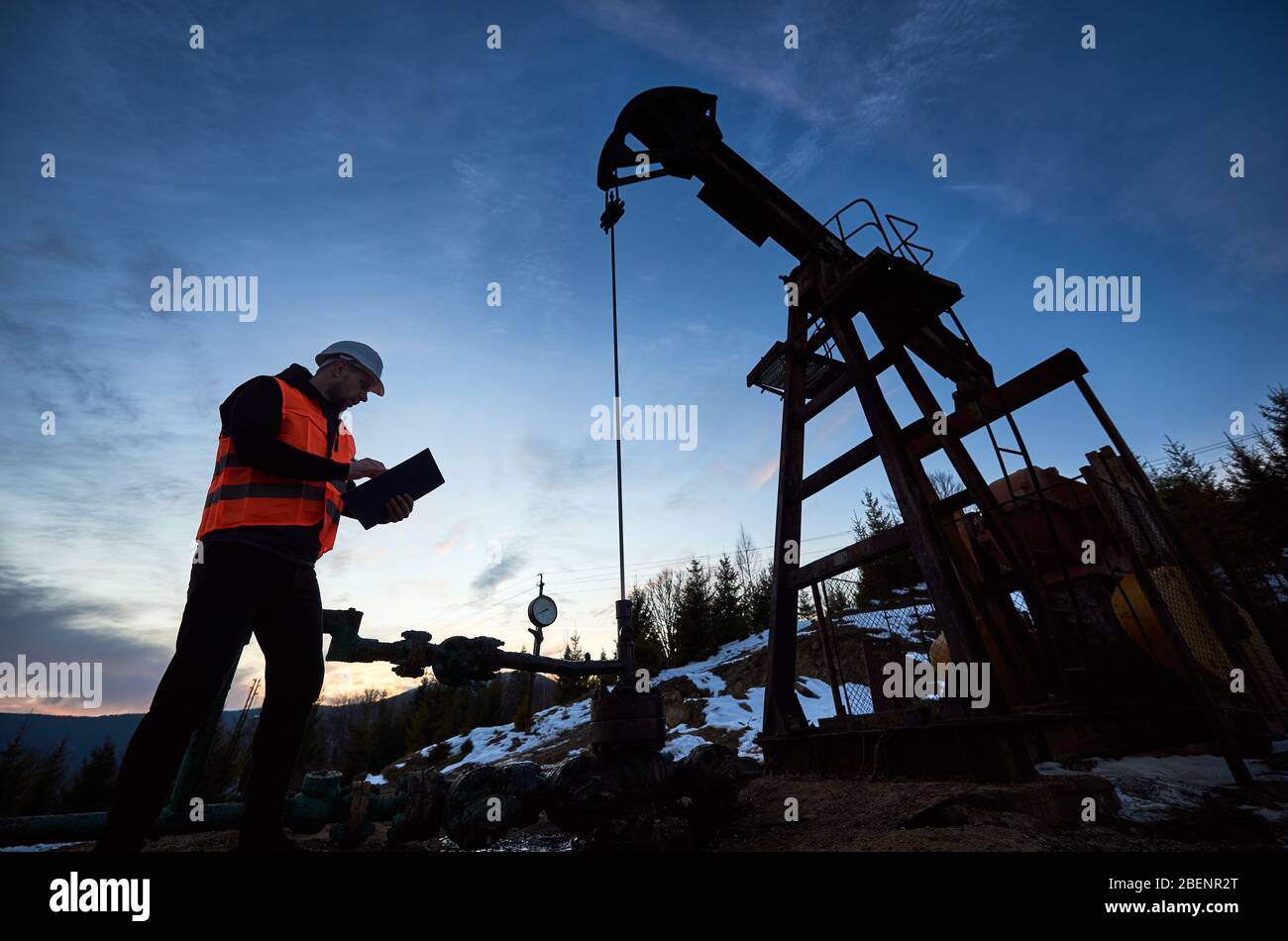 Oil engineer in work vest at oil pump rocker-machine, making notes ...