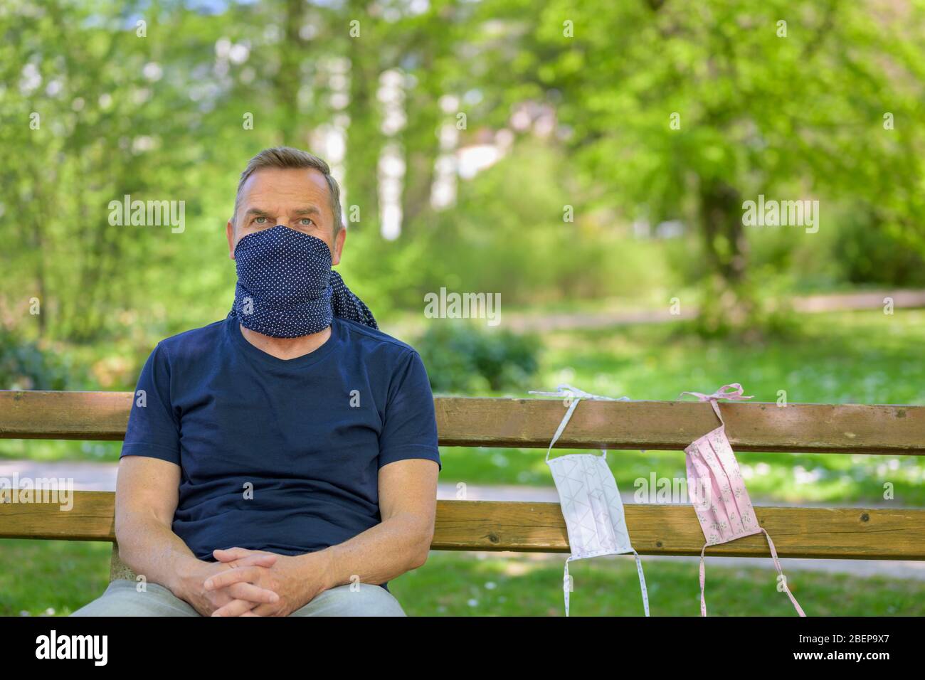 Man wearing a face guard seated on a park bench with two surgical masks ...