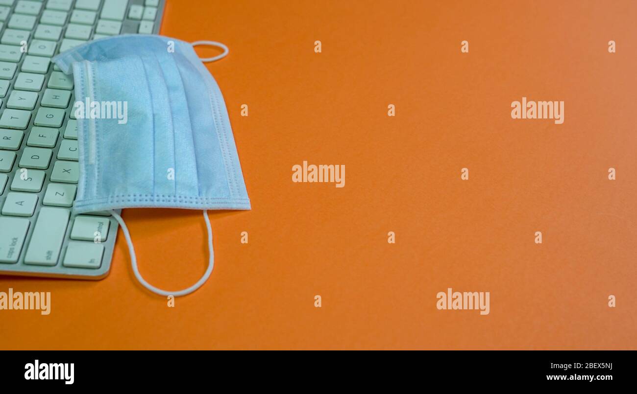 Medical face mask on top of keyboard. Orange background. Top view. Copy ...