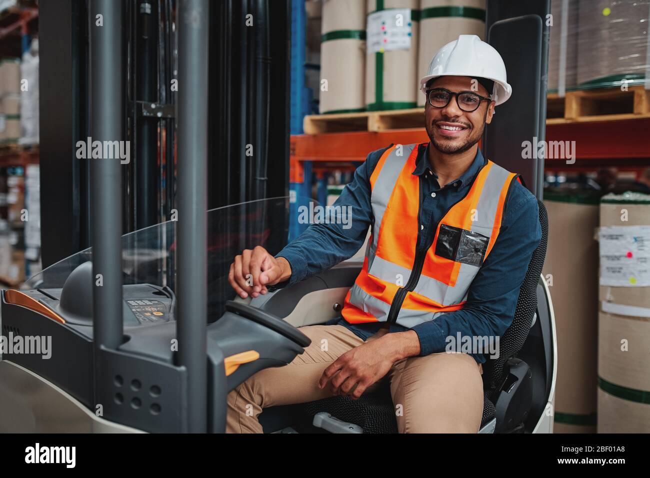 Smiling forklift driver with spectacles sitting in vehicle in warehouse ...