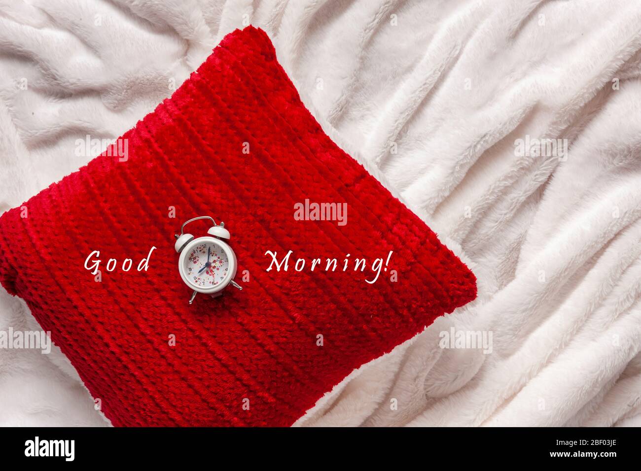 Good Morning. Close up of a red pillow and alarm clock on white blanket ...