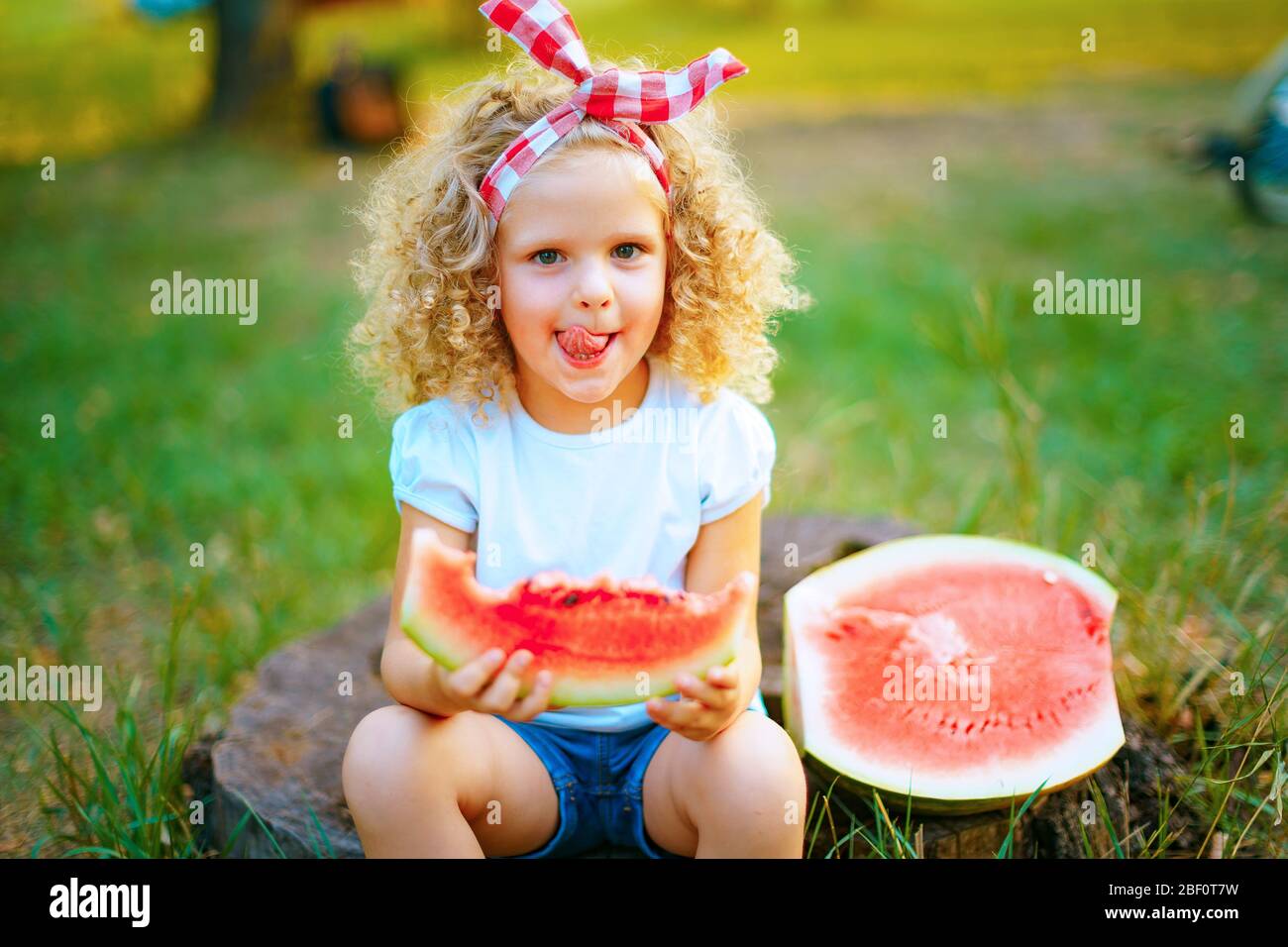 Happy curly child girl sitting on stump and eating watermelon outdoors ...