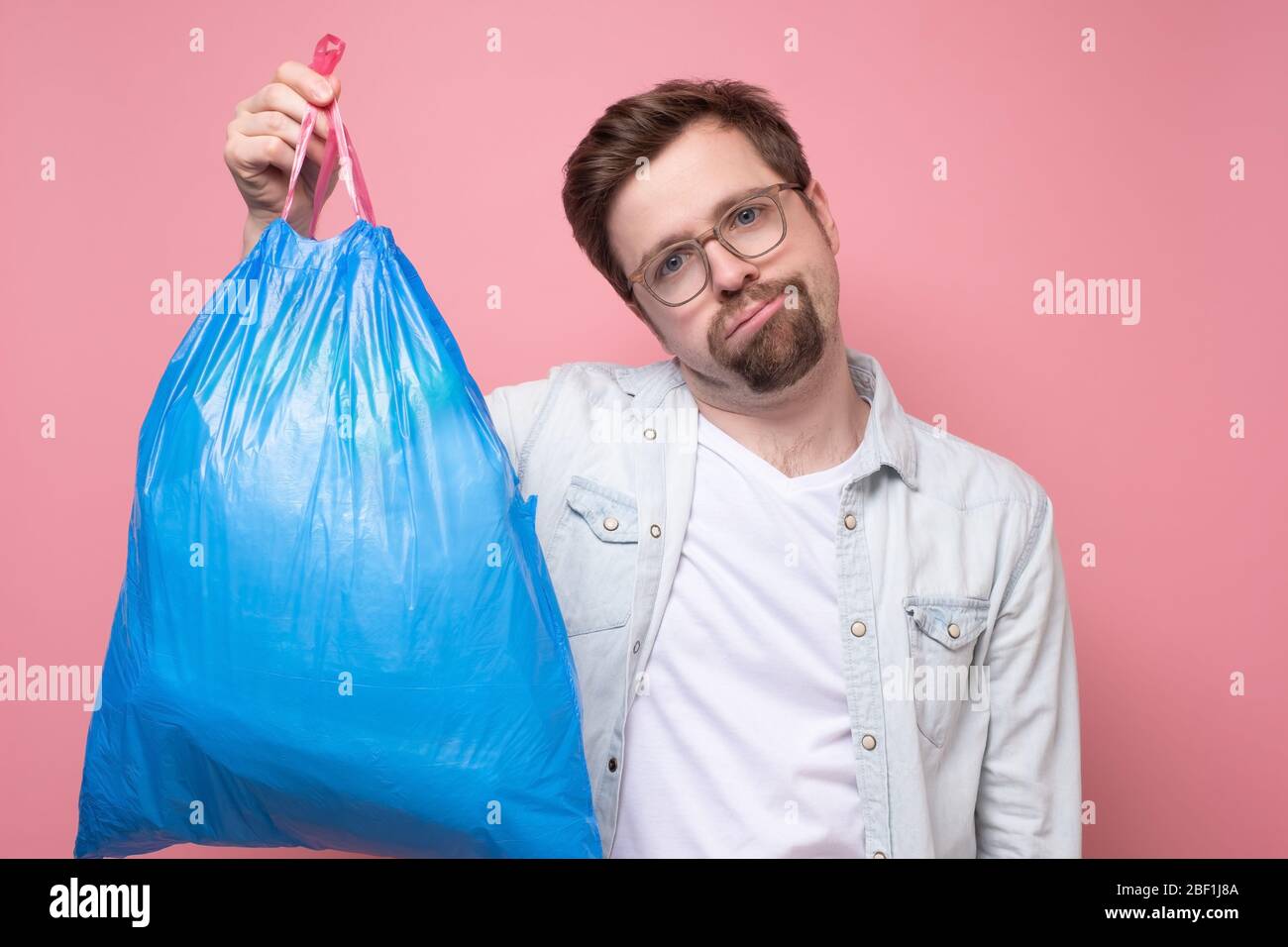 Handsome disappointed man holding blue garbage bag isolated on pink ...