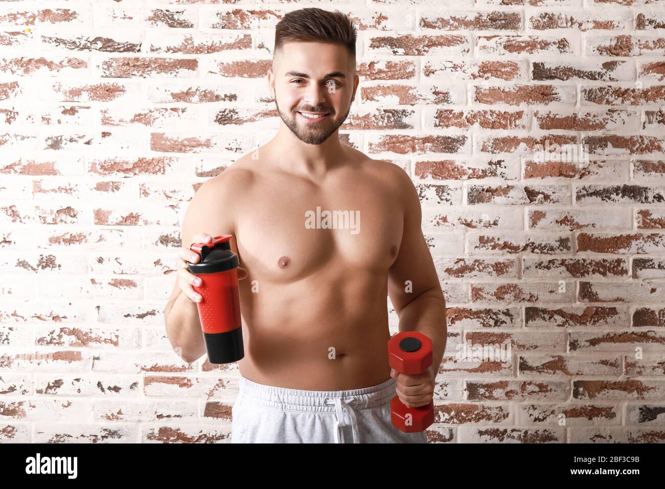Sporty man with protein shake and dumbbell on brick background Stock ...