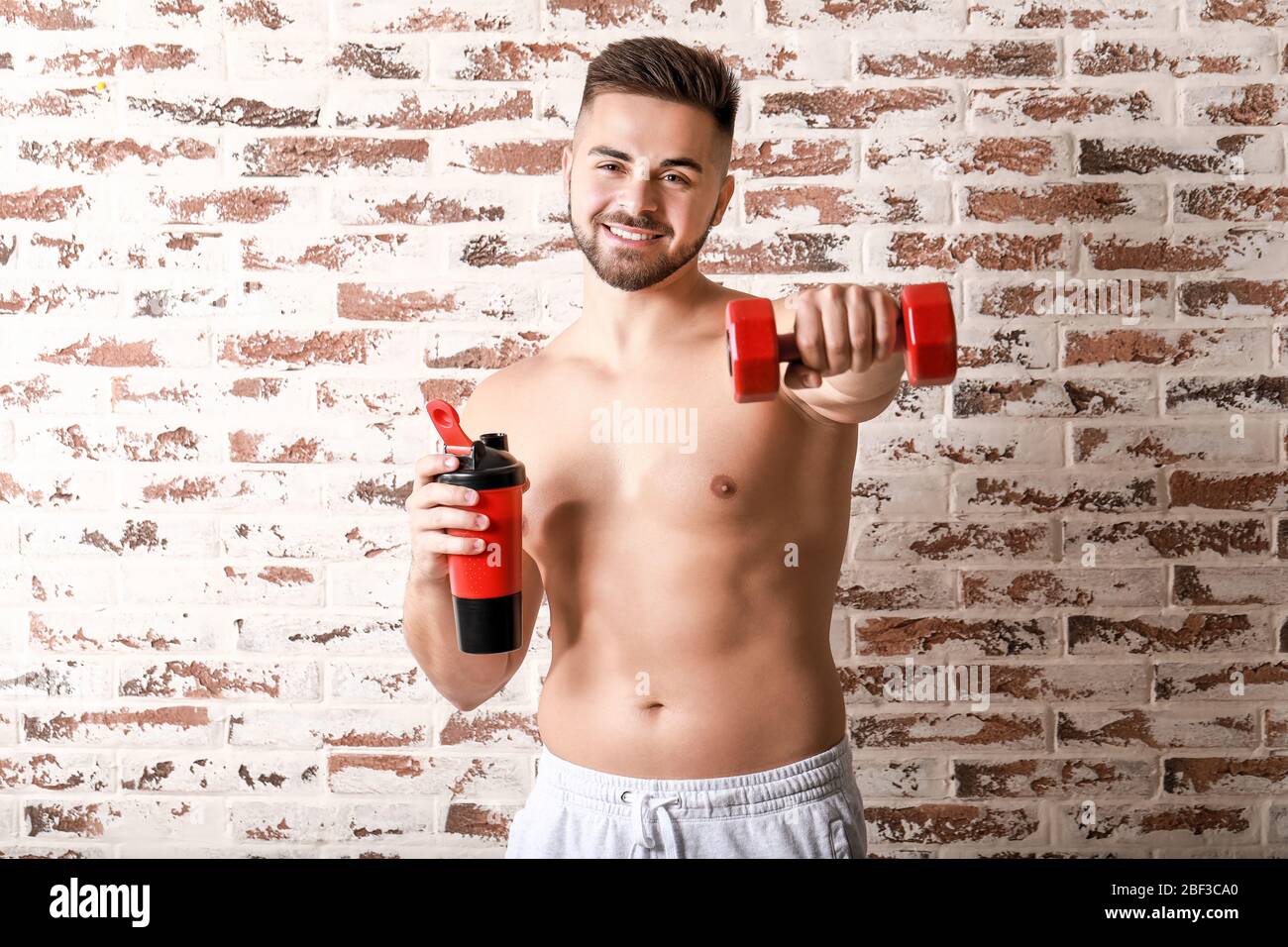 Sporty man with protein shake and dumbbell on brick background Stock ...