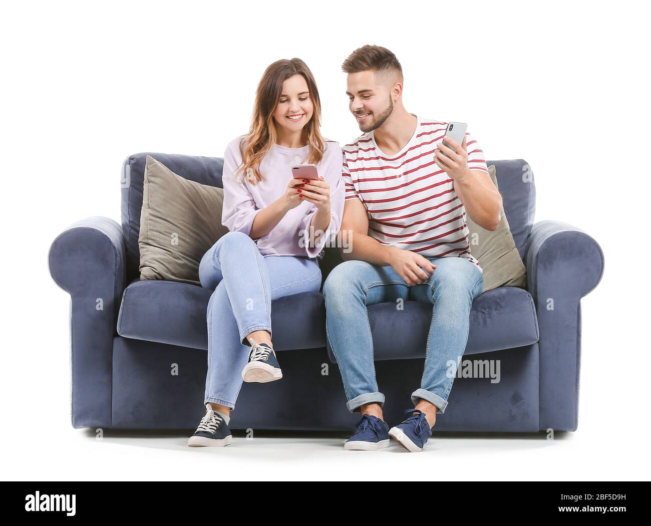 Young couple with mobile phones sitting on sofa against white ...