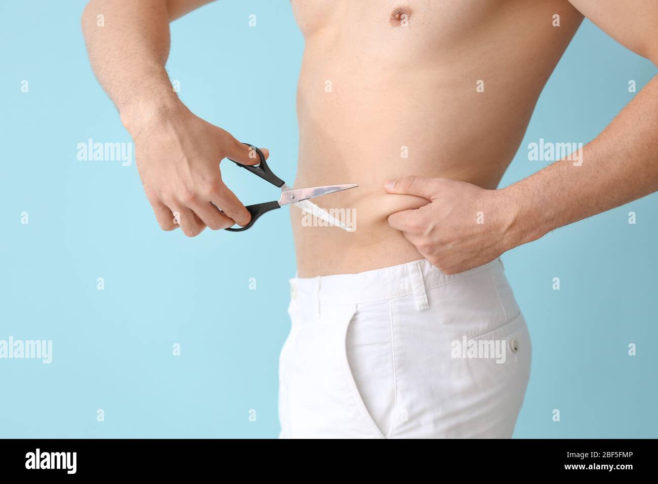 Young man with scissors touching his belly fat on color background ...