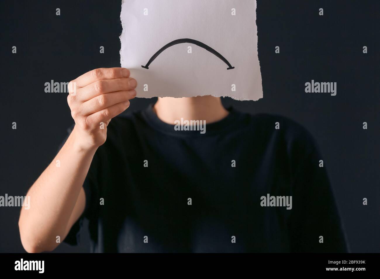 Woman holding paper sheet with drawn sad mouth on dark background ...