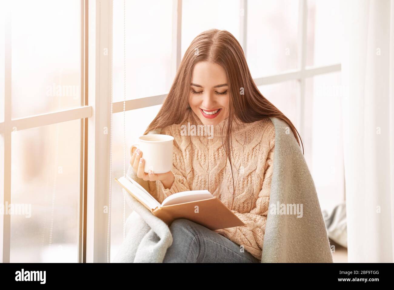 Beautiful young woman drinking hot cocoa and reading book near window ...