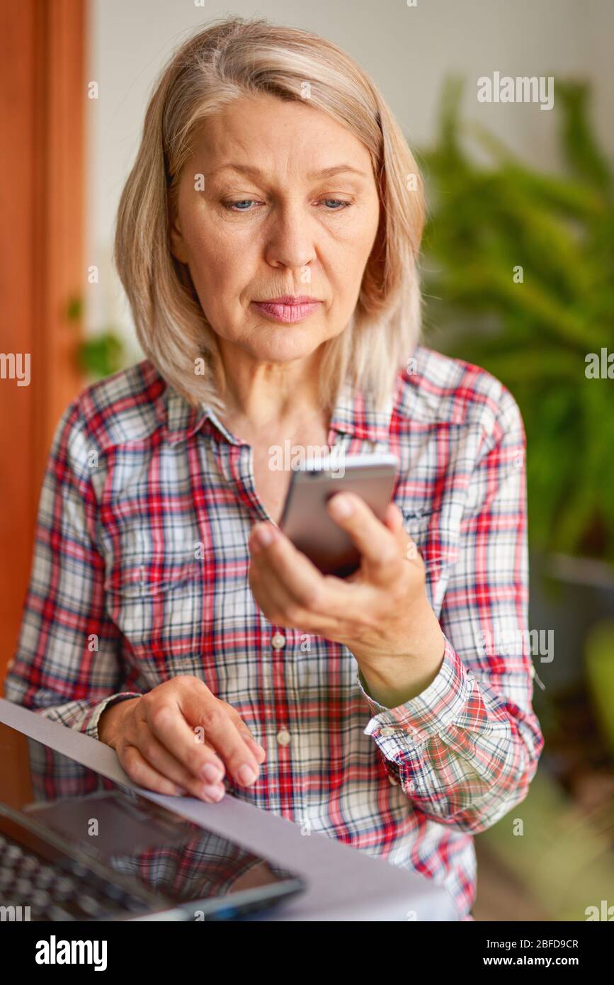 Mature woman with a phone in her hand looking at laptop at home Stock ...