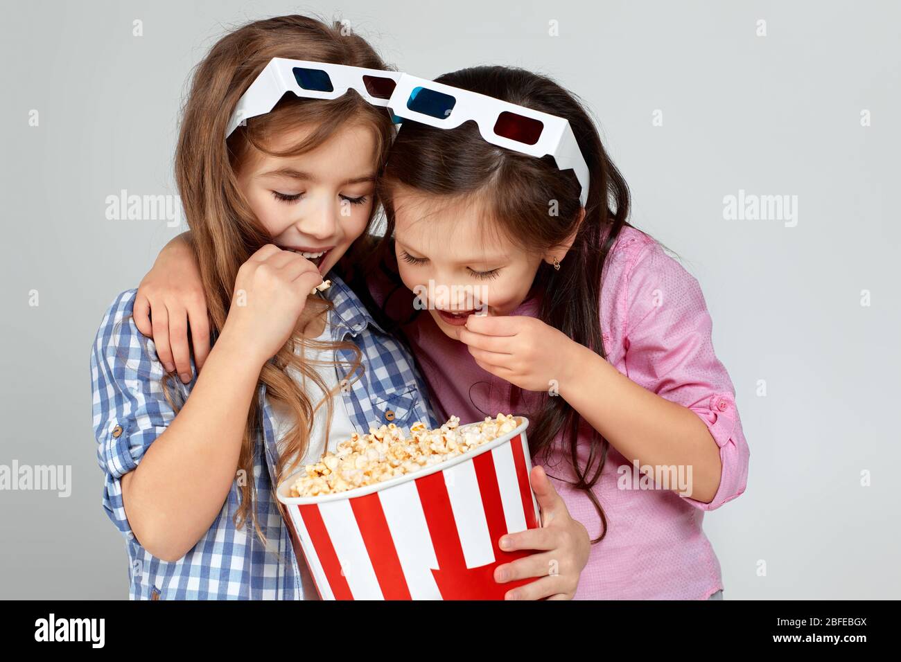 two beautiful caucasian little girls wearing red-blue 3d glasses and eating popcorn from bucket ...
