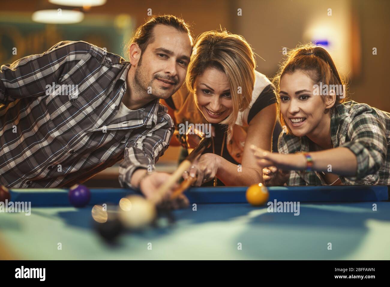 Group of a smiling cheerful friends are playing billiards in bar after ...