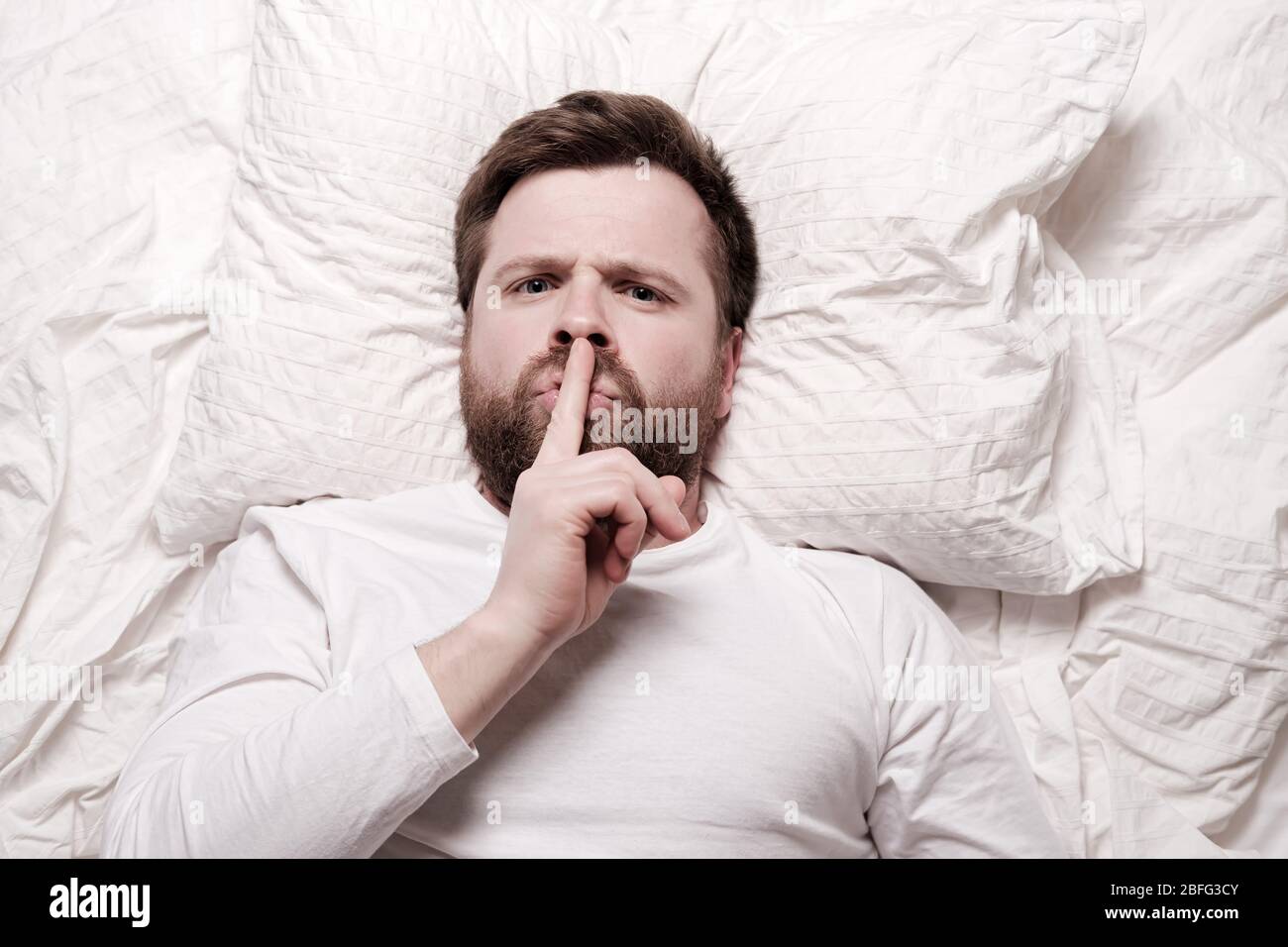 Man is lying in bed and puts index finger to lips, expressing silence ...