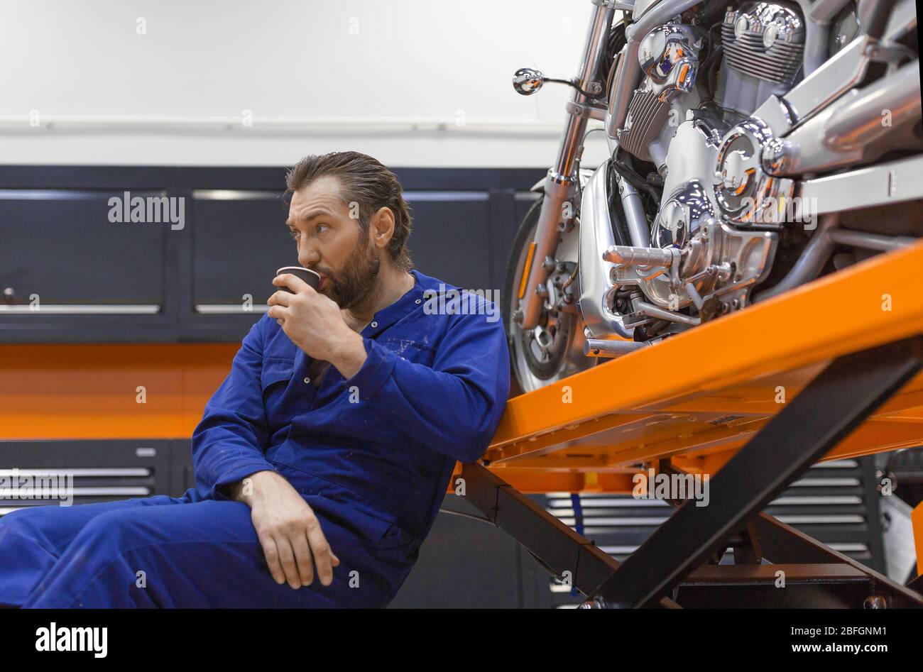 A man in the workshop sits near a motorcycle and drinking coffee. Rest ...
