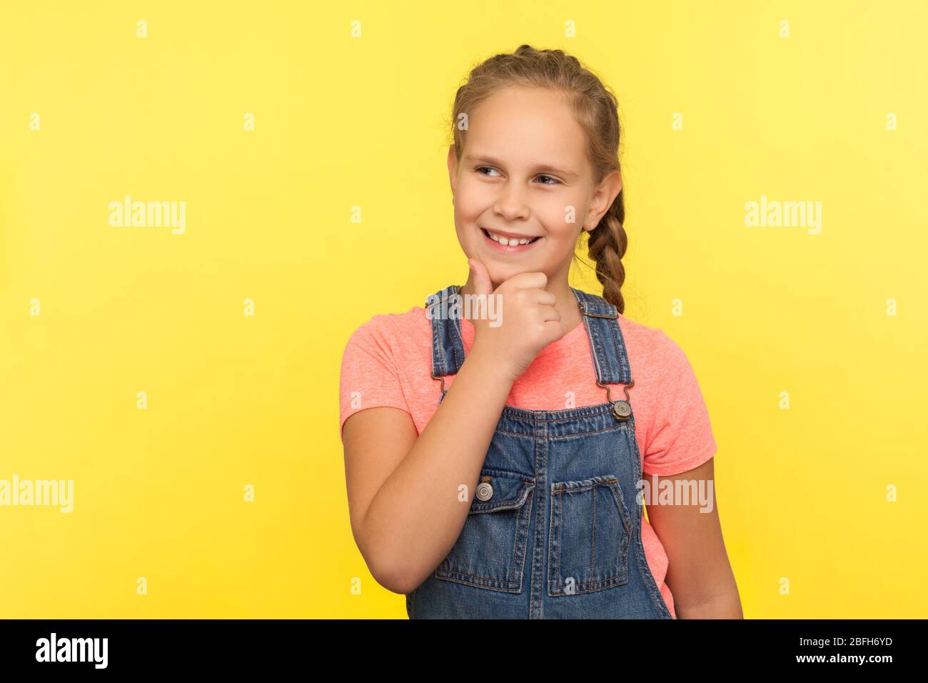 Portrait of positive thoughtful little girl in denim overalls holding ...