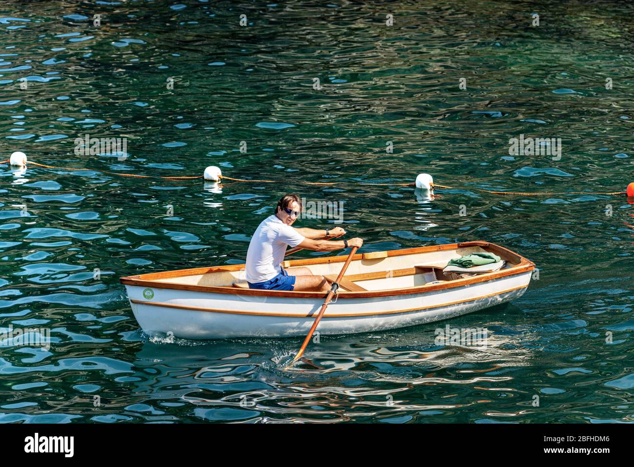 A man on a small wooden rowboat in the blue Mediterranean sea in front ...