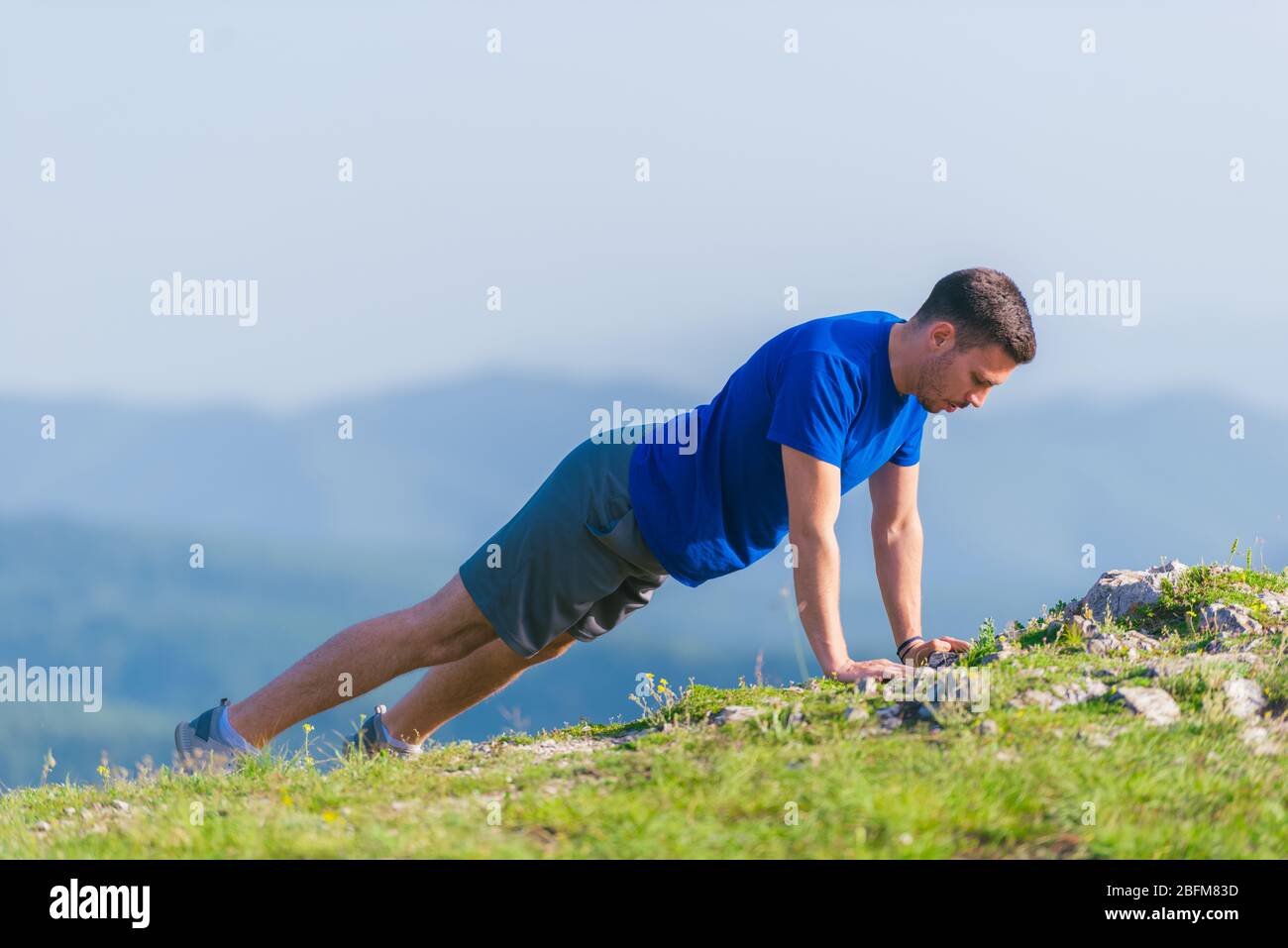 A young fit male athlete is doing push-ups outdoors on a cliff while ...
