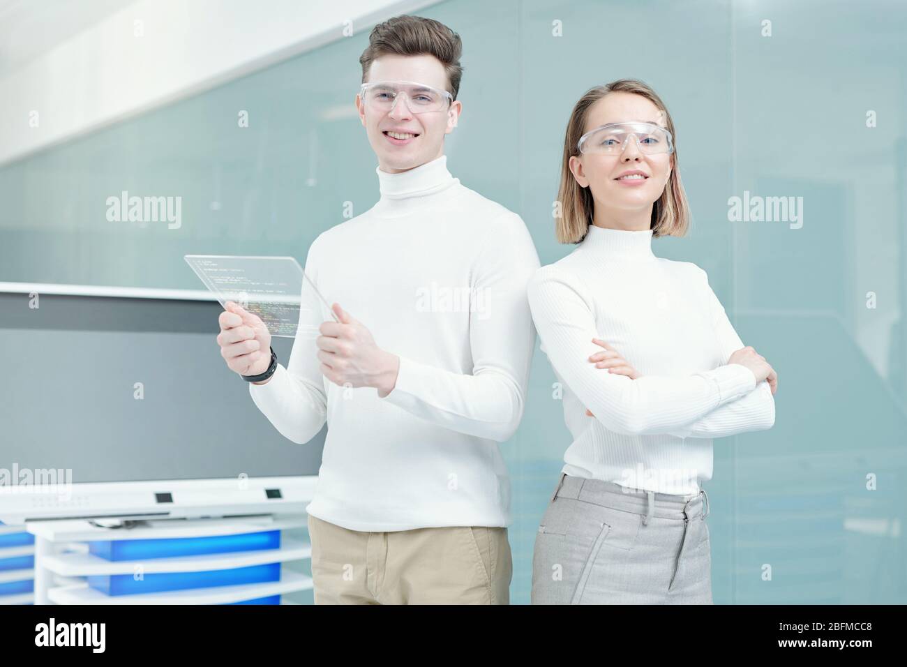 Two young smiling programmers in smart casualwear and goggles standing ...