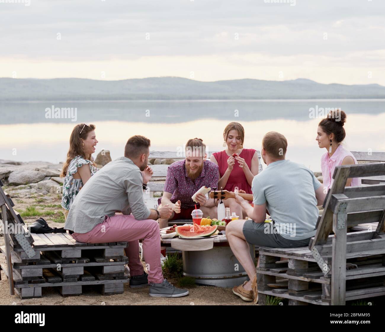 Group of friends sitting around table near picturesque lake on summer ...