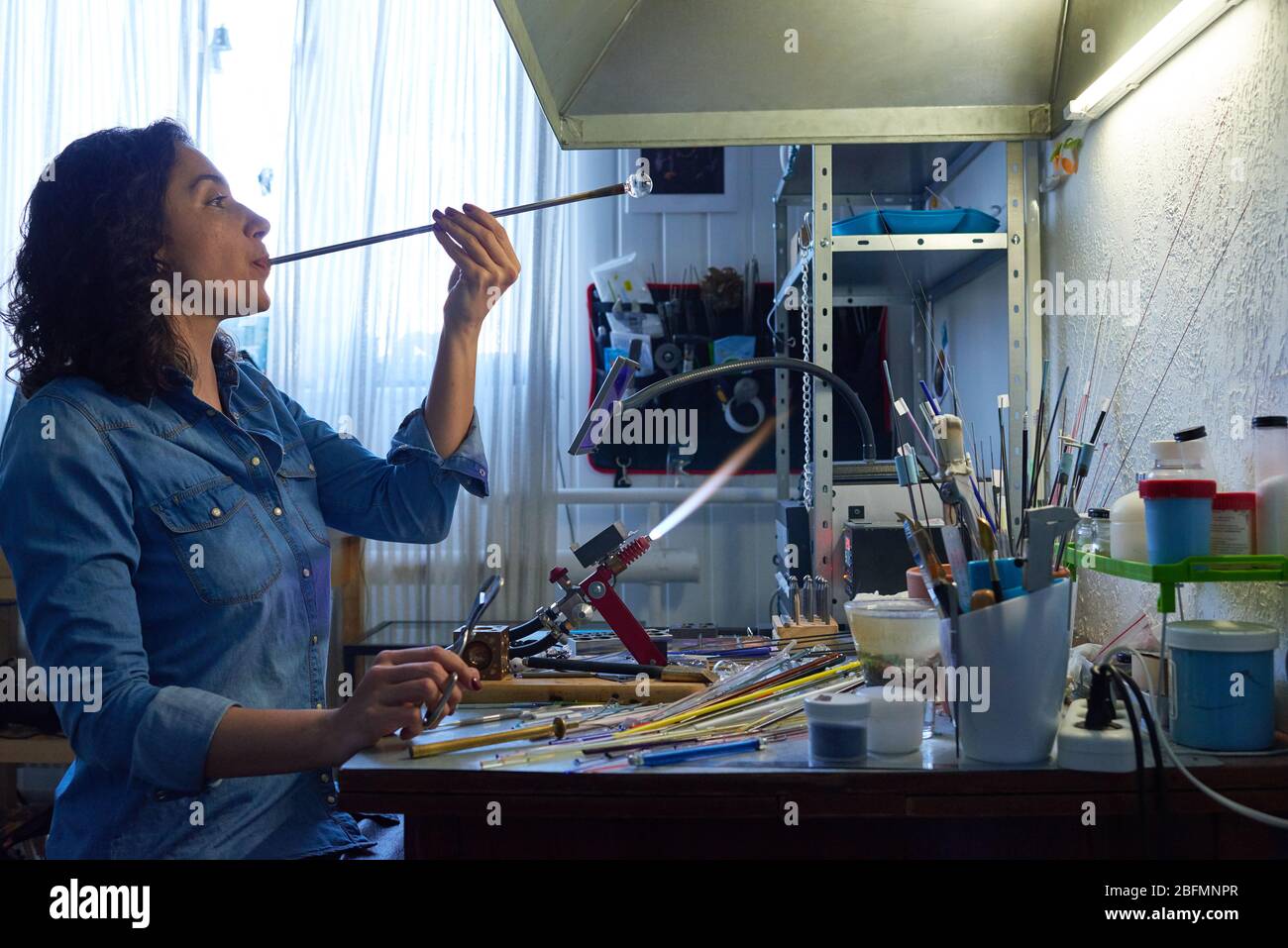 Young skilled female lampworking master blowing into long tube while ...