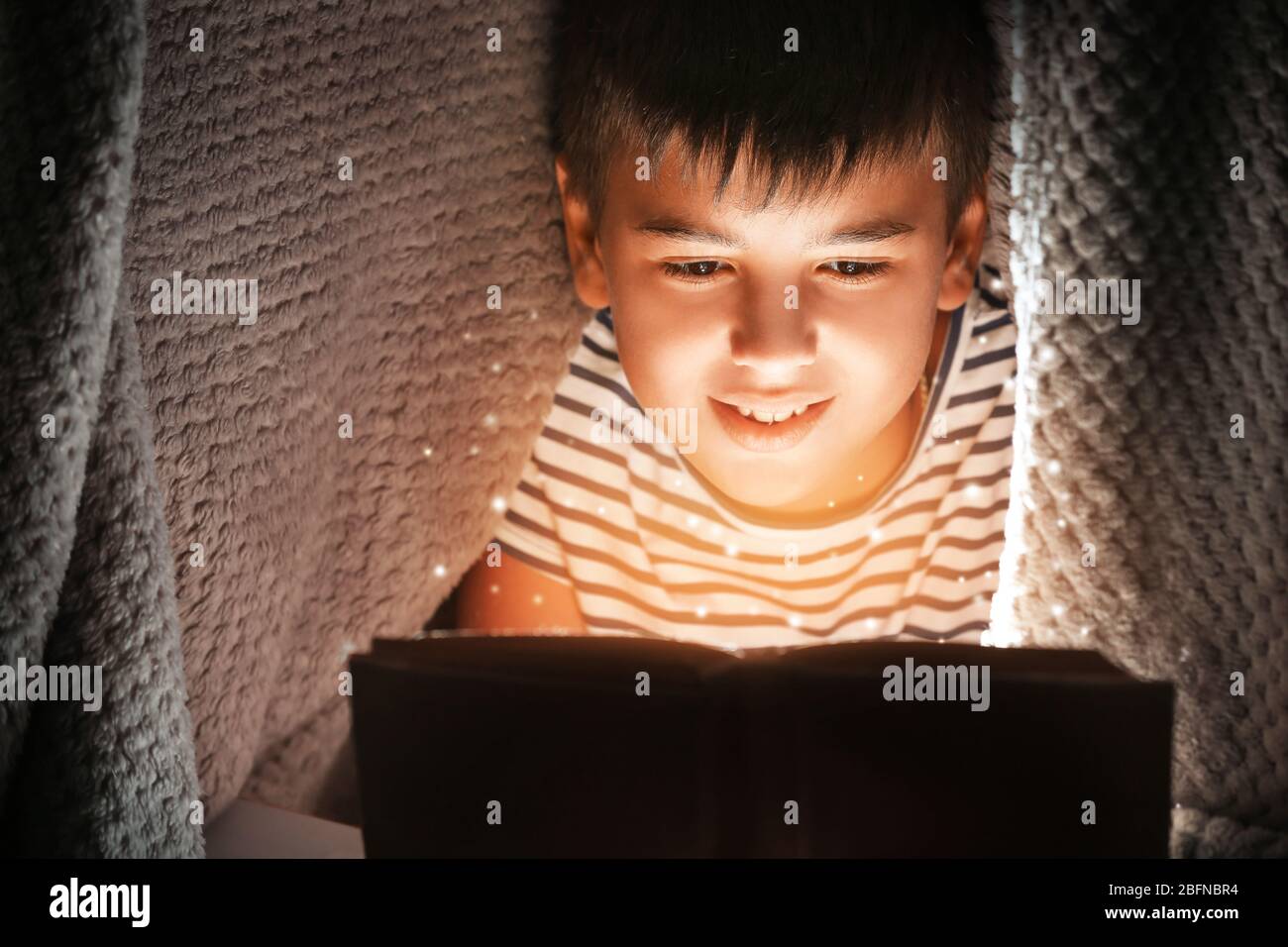 Cute boy reading book under blanket at night. Magic light coming out of ...