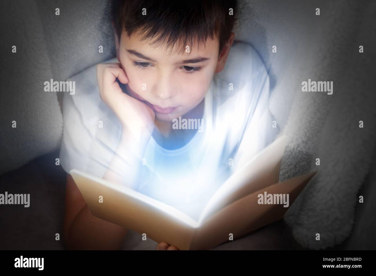Cute boy reading book under blanket at night. Magic light coming out of ...