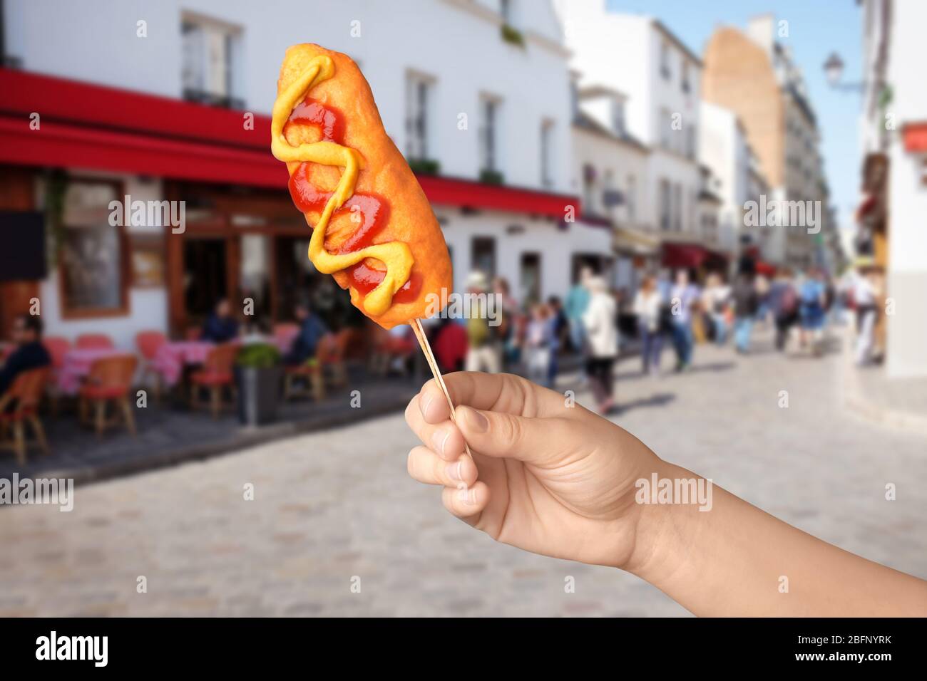Woman holding tasty corn dog with ketchup and mustard on city street ...