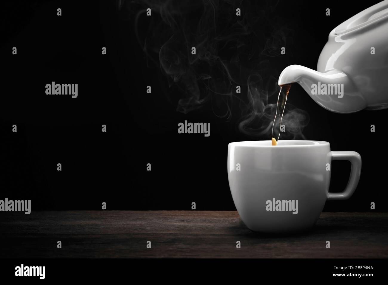 Pouring hot tea from a kettle into a cup on black background, close up ...