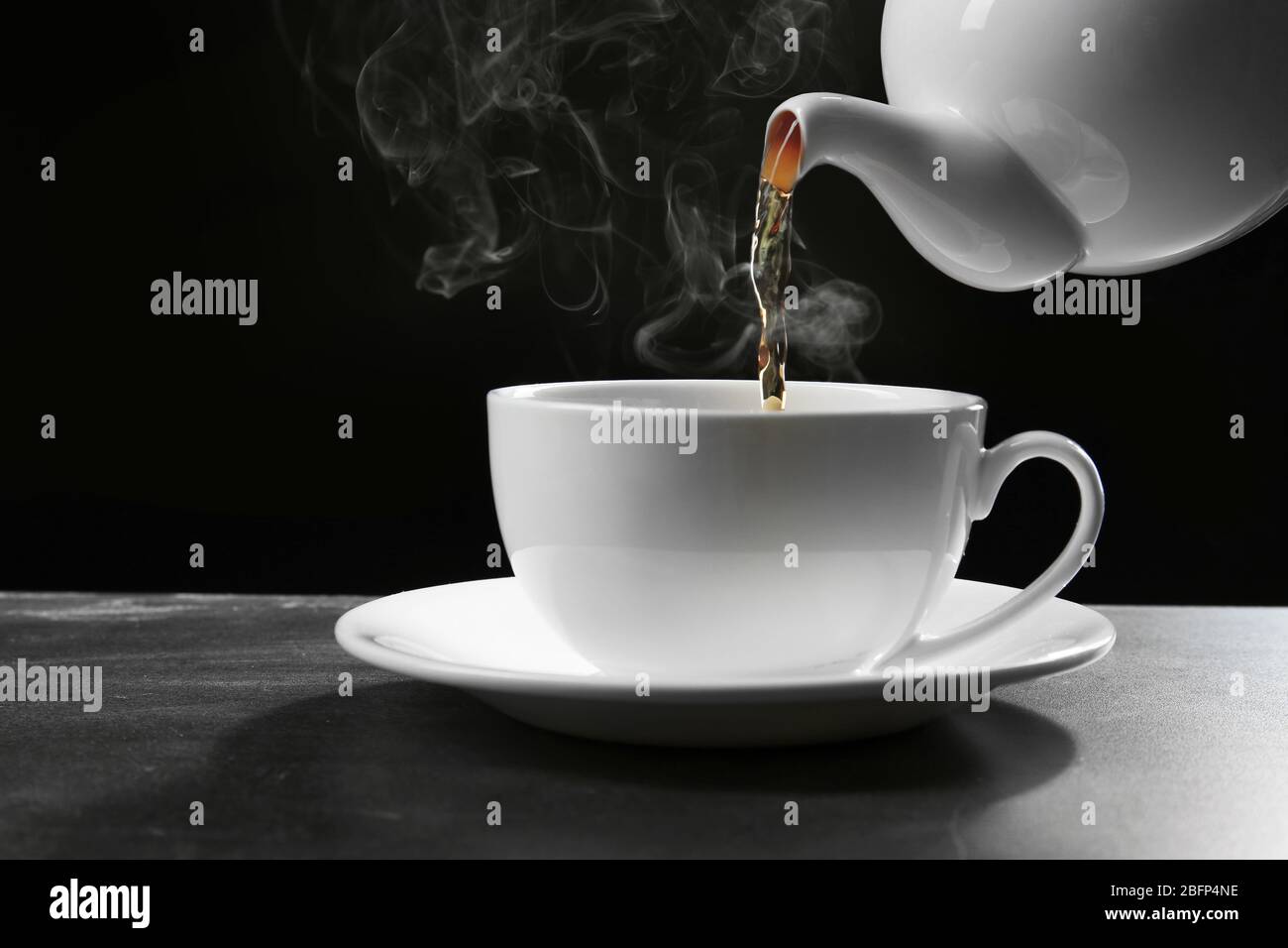 Pouring hot tea from a kettle into a cup on black background, close ...