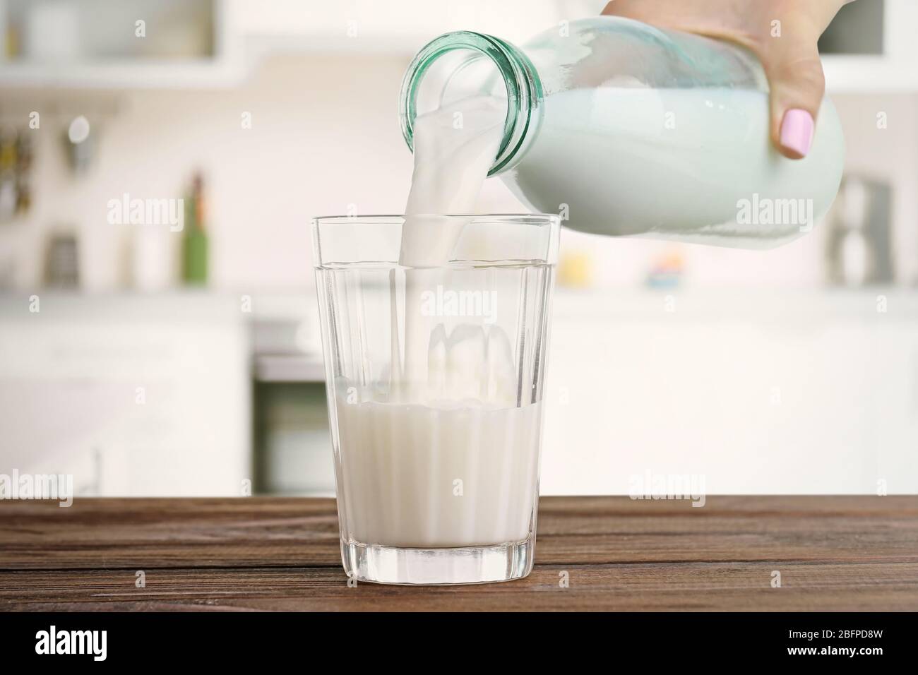 Female hand pouring milk into glass against blurred kitchen background ...