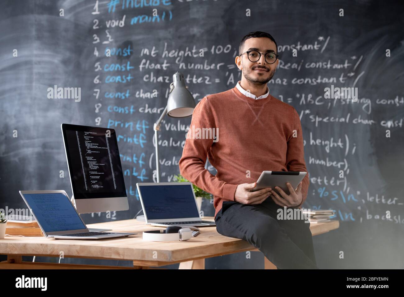 Portrait of confident young Arabian coder in glasses sitting on table and using tablet against ...