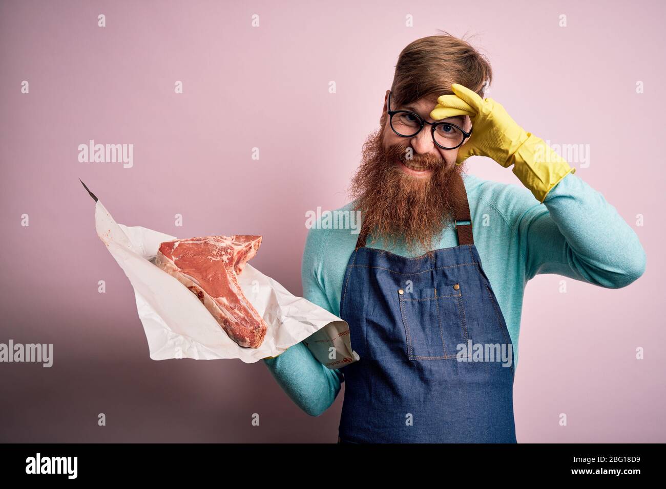 Redhead Irish butcher man with beard holding raw beef steak over pink ...