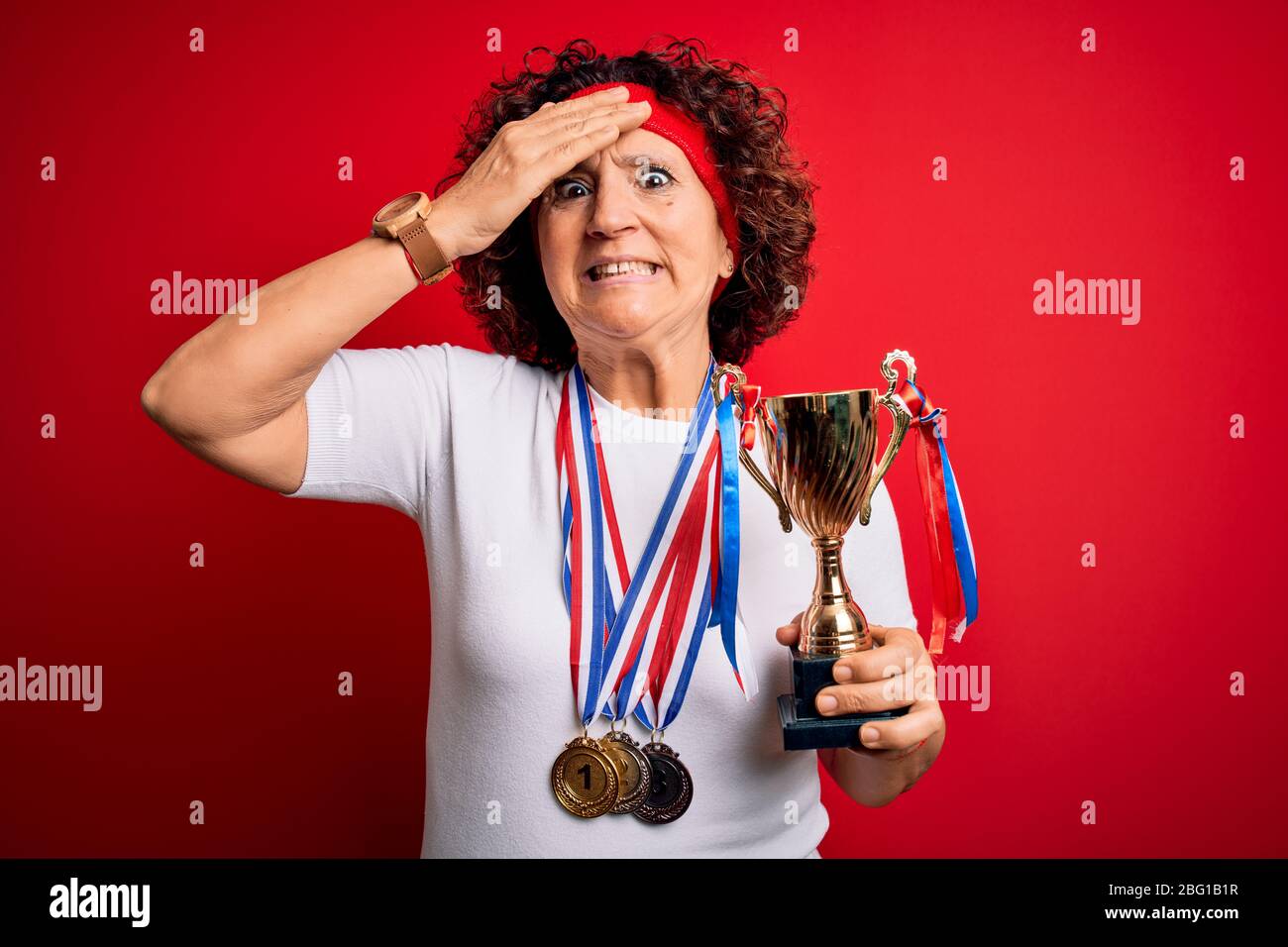 Middle age curly woman winning medals holding trophy over isolated red ...