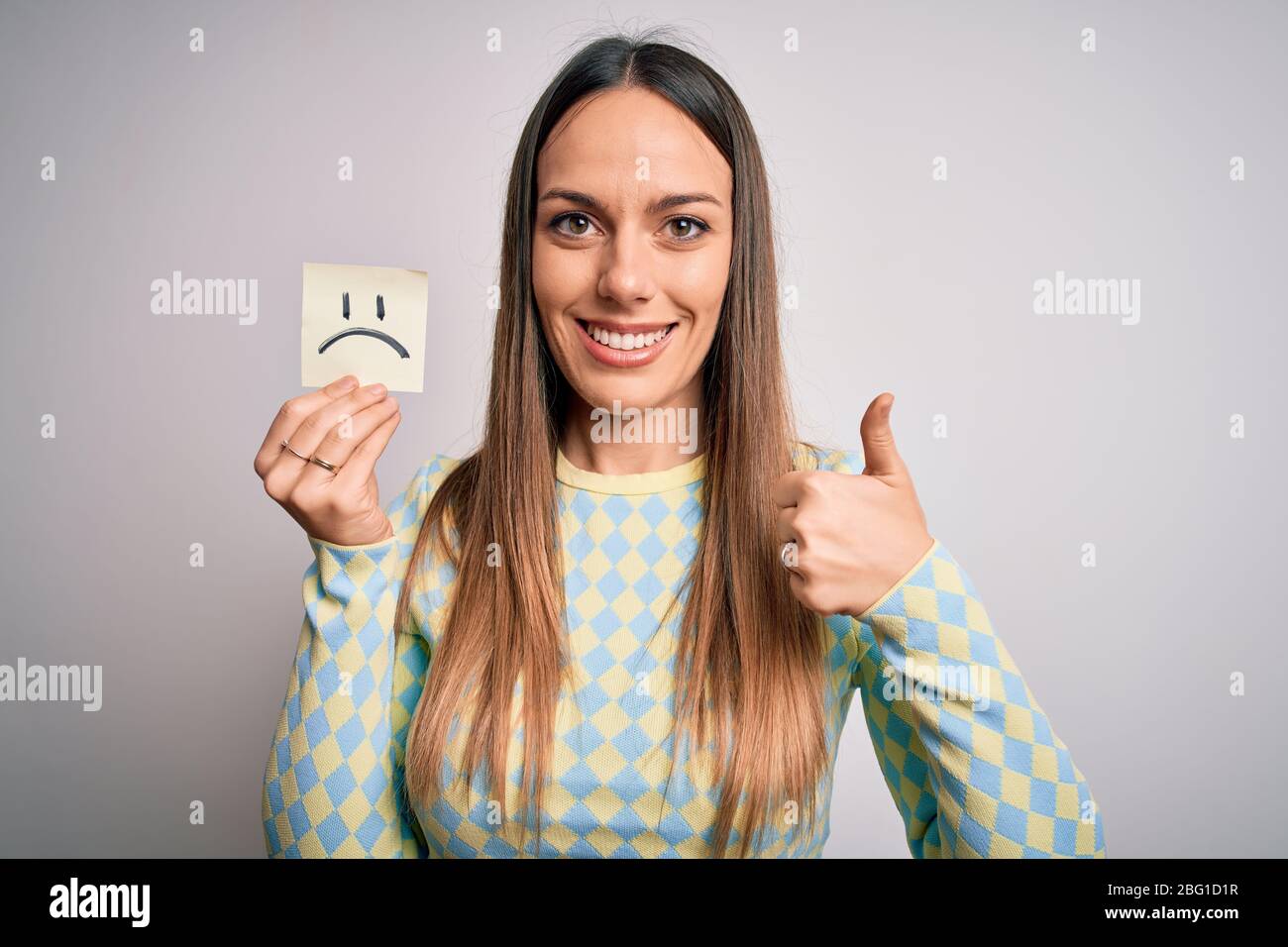 Young blonde woman holding sticky paper note with sad face emoticon ...