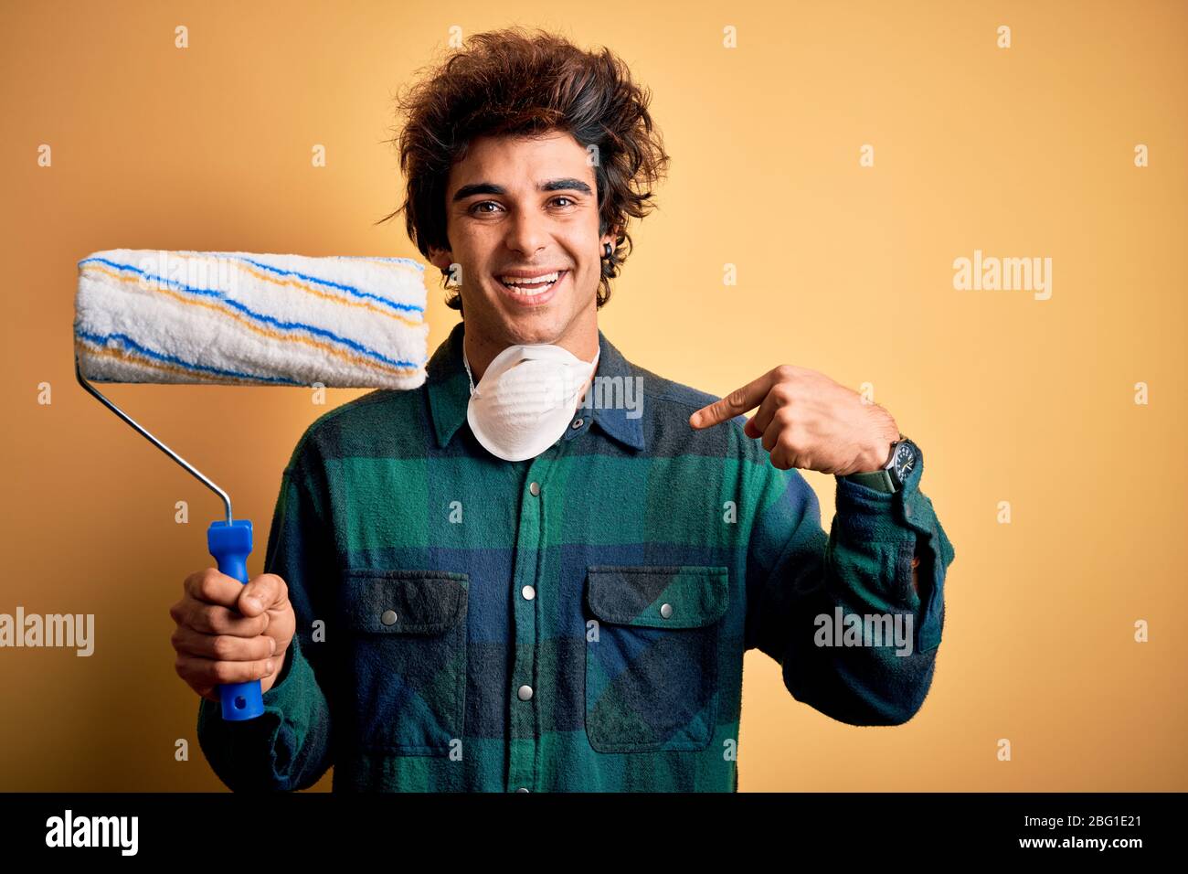 Young handsome painter man holding roller standing over isolated yellow ...