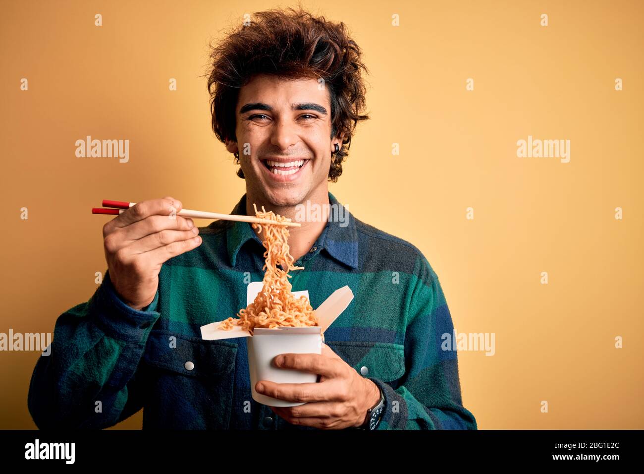 Young handsome man eating noodles standing over isolated yellow ...
