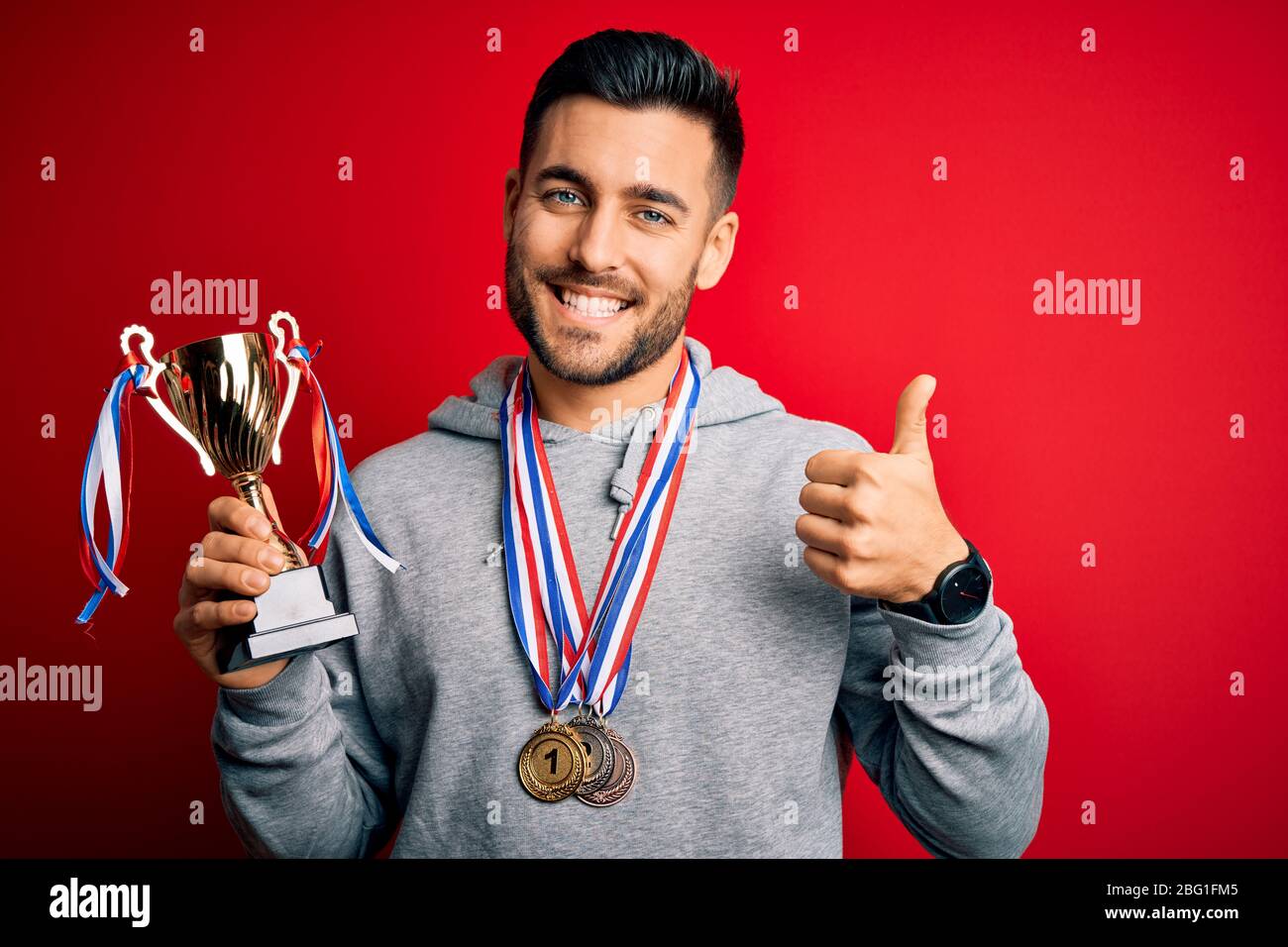 Young handsome succesful man holding trophy wearing medals over red ...