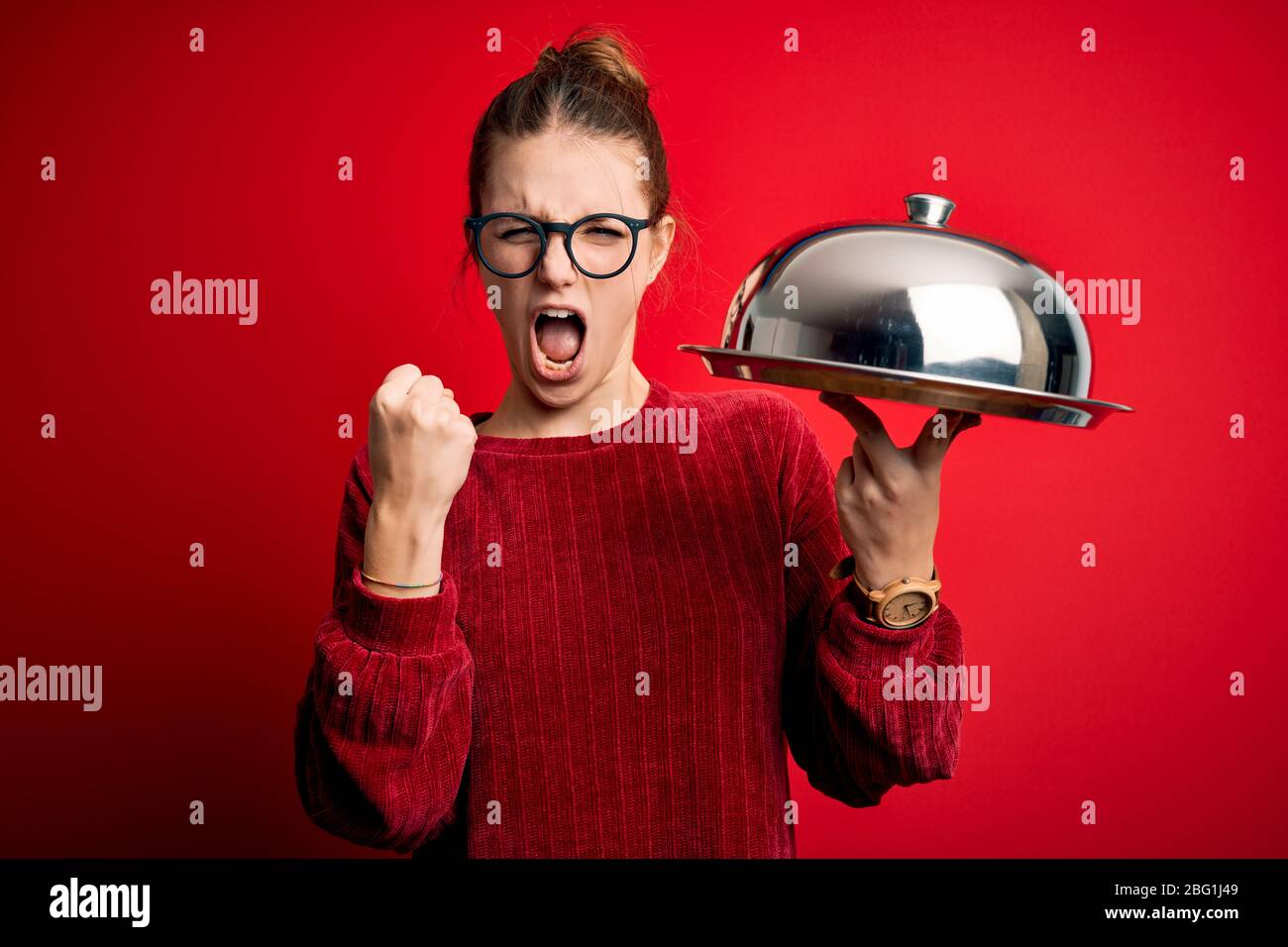 Young beautiful redhead woman holding waitress tray over isolated red ...