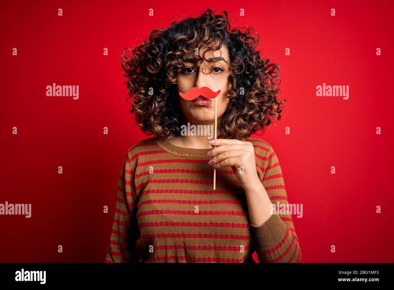Young beautiful curly arab woman holding funny mustache over isolated ...