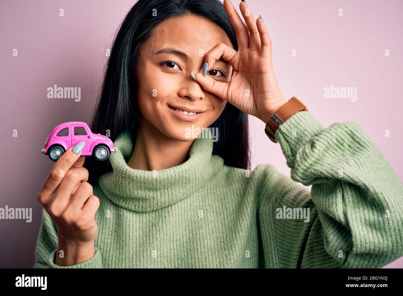 Young beautiful chinese woman holding small toy car over isolated pink ...