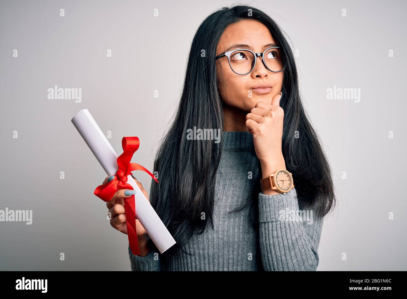 Young beautiful chinese woman holding diploma standing over isolated white background serious ...