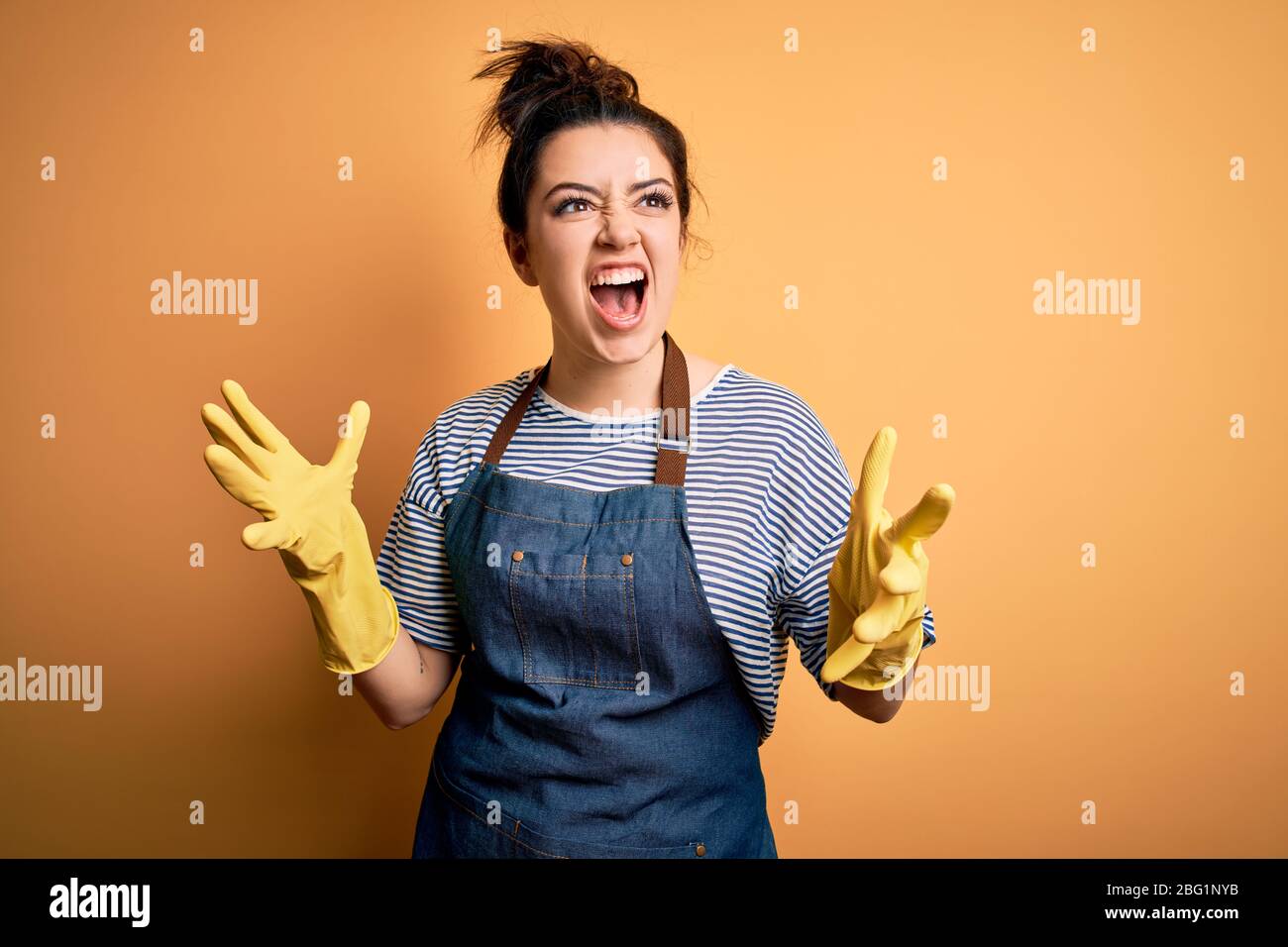 Young brunette cleaner woman wearing housekeeping gloves over yellow ...