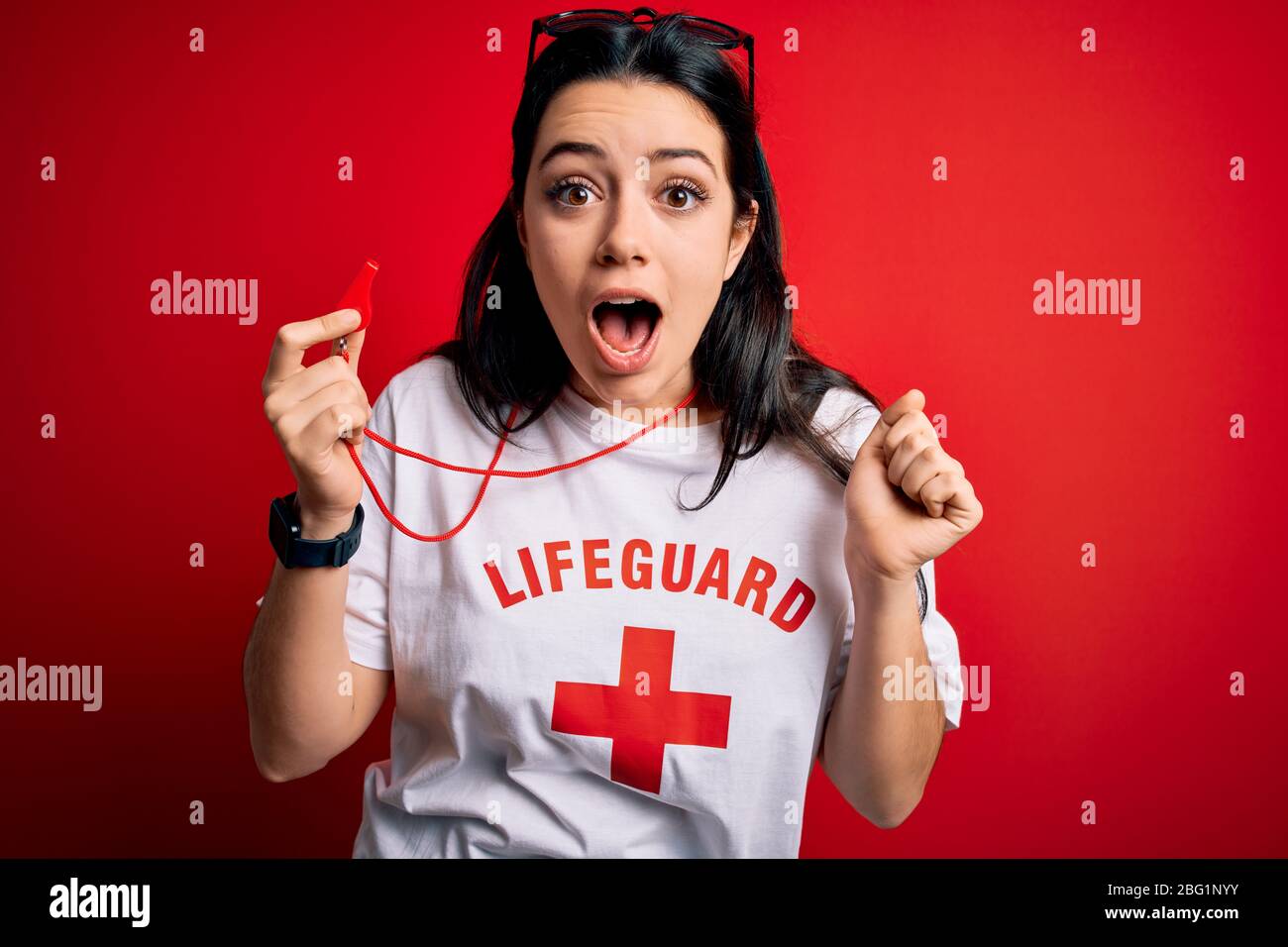 Young lifeguard woman wearing guard equipement holding whistle over red ...