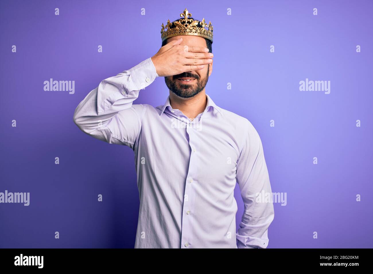 Young handsome man with beard wearing golden crown of king over purple ...