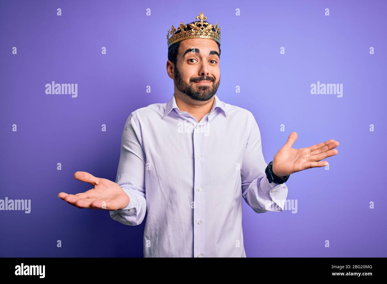 Young handsome man with beard wearing golden crown of king over purple ...