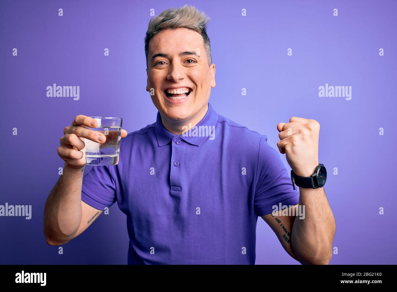 Young modern man drinking a glass of fresh water over purple isolated ...